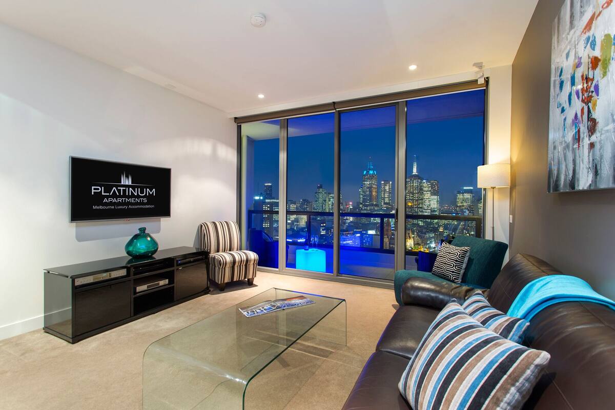 Uma sala de estar moderna com uma grande janela que oferece vista para a cidade, uma televisão de tela plana na parede, um sofá de couro e uma mesa de centro de vidro.