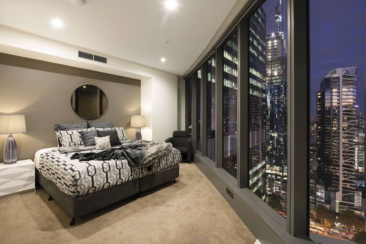 Quarto moderno com cama grande, duas luminárias de cabeceira, um espelho redondo e vista para prédios da cidade à noite.