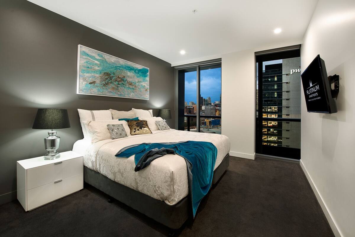 Um quarto moderno com uma cama grande, uma mesa de cabeceira, um quadro e vista da cidade por uma porta de vidro.