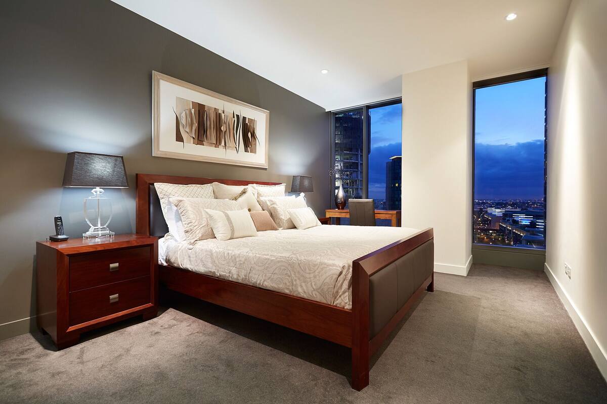 Quarto moderno com uma cama grande, mesa de cabeceira, escrivaninha e vista da cidade através de uma janela ampla.