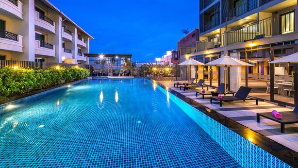 Piscina com espreguiçadeiras e guarda-sóis, cercada por edifícios com varandas e áreas de estar ao ar livre.
