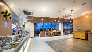 Um lobby de hotel moderno com área de buffet, piso de madeira e grandes janelas com vista para a paisagem urbana.