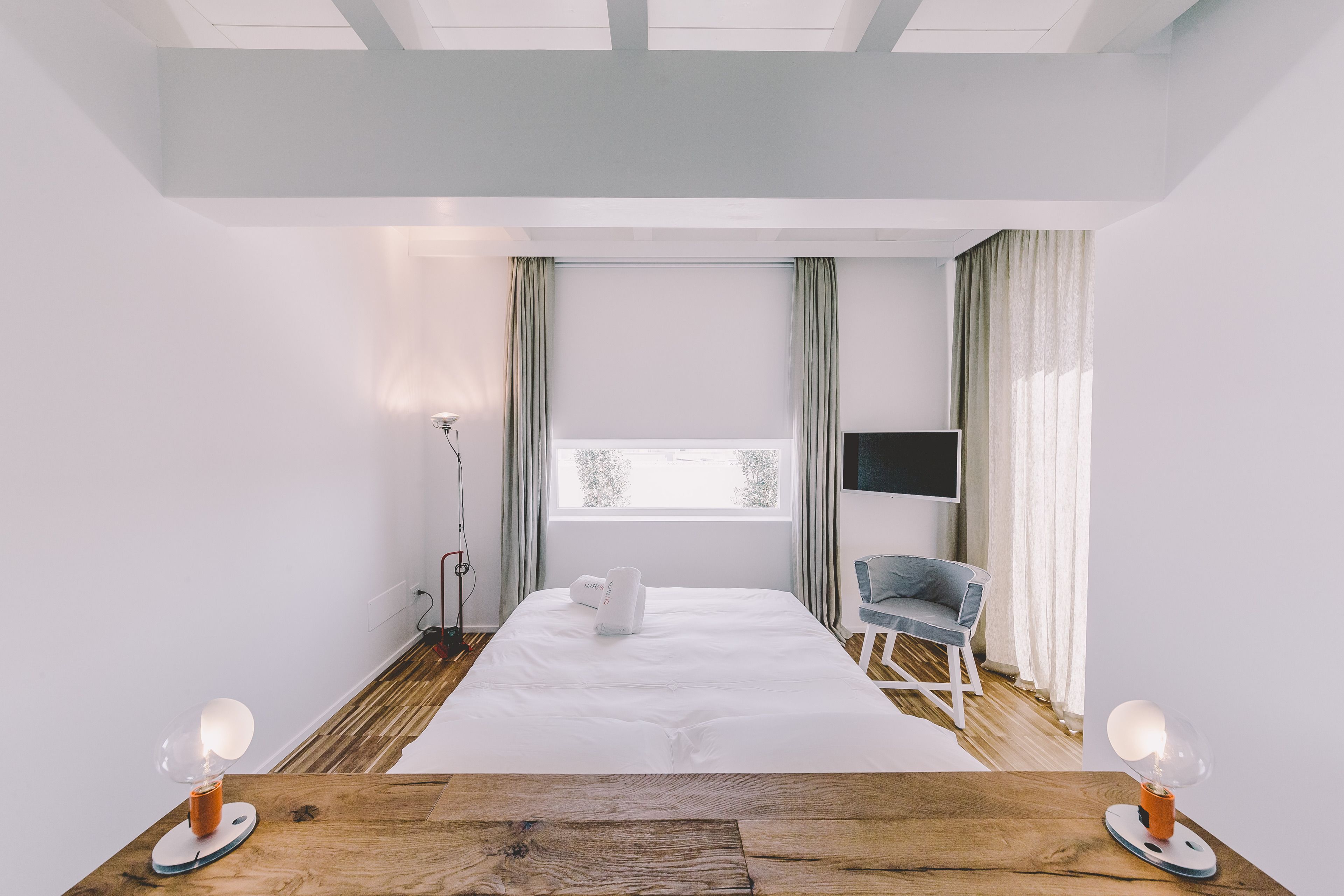 Quarto de hotel moderno com uma cama grande, uma escrivaninha com abajur, uma cadeira e uma TV na parede.