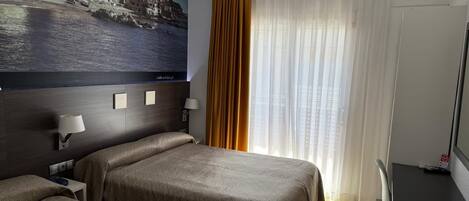 Quarto de hotel com duas camas, uma janela grande com cortinas, uma televisão fixada na parede e um quadro de uma vila costeira.