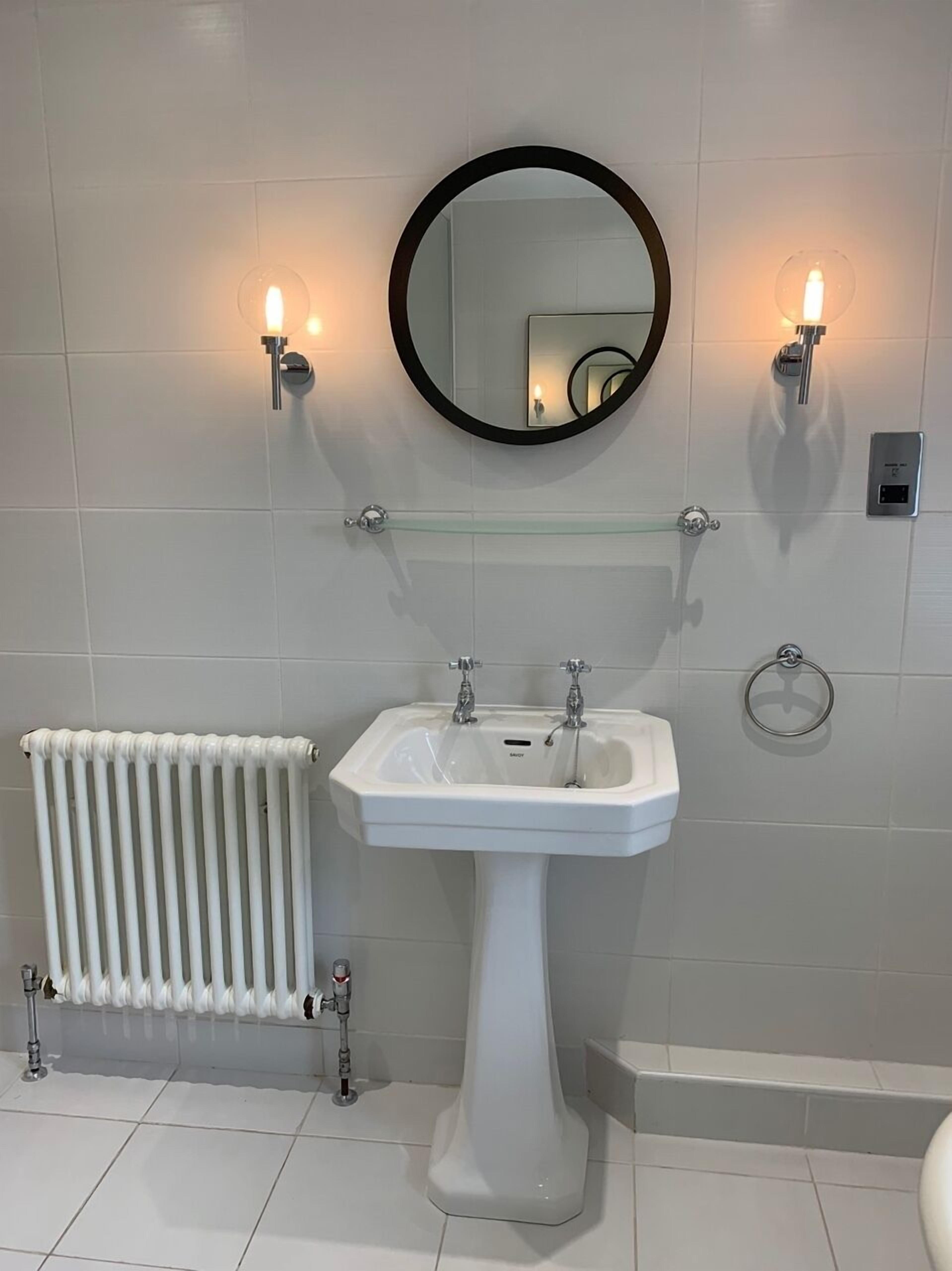 Uma casa de banho com lavatório assentado em pedestal, duas luzes fixadas na parede, um espelho redondo, uma prateleira de vidro, um suporte para toalhas e um radiador branco.