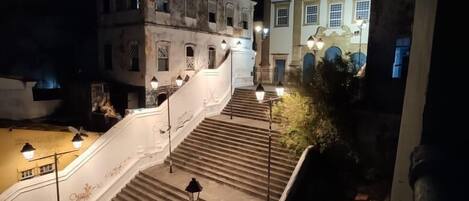 Vista noturna de um edifício histórico com escadaria que leva a um segundo andar, cercado por outros edifícios com janelas iluminadas.