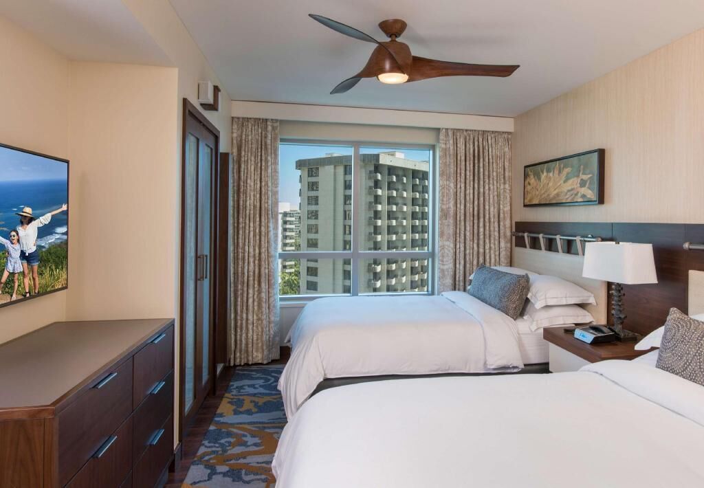 Quarto de hotel com cama, escrivaninha, ventoinha no teto, janela com cortinas e um quadro na parede.