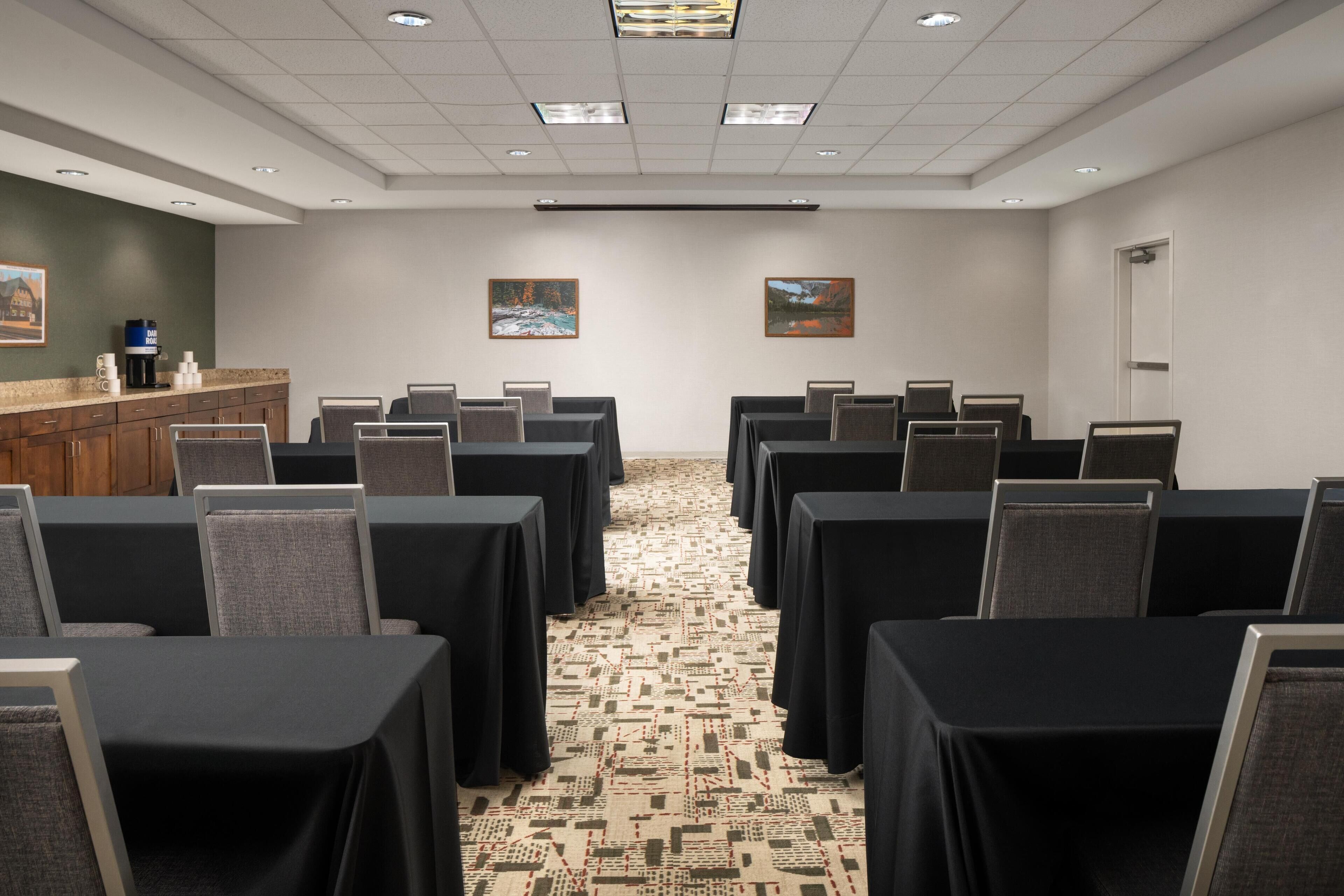 Uma sala de conferências com mesas e cadeiras dispostas em fileiras, um espaço para café e duas pinturas nas paredes.