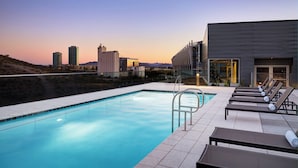 Uma área de piscina moderna com um horizonte urbano ao fundo, cadeiras de descanso e céu claro.