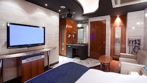 Quarto de hotel moderno com televisão de ecrã plano, casa de banho pequena com lavatório e espelho, cama com colcha azul e área de estar com uma cadeira e uma mesa pequena.
