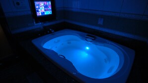 Um quarto com pouca luz, uma banheira grande, televisão na parede e paredes revestidas com azulejos.