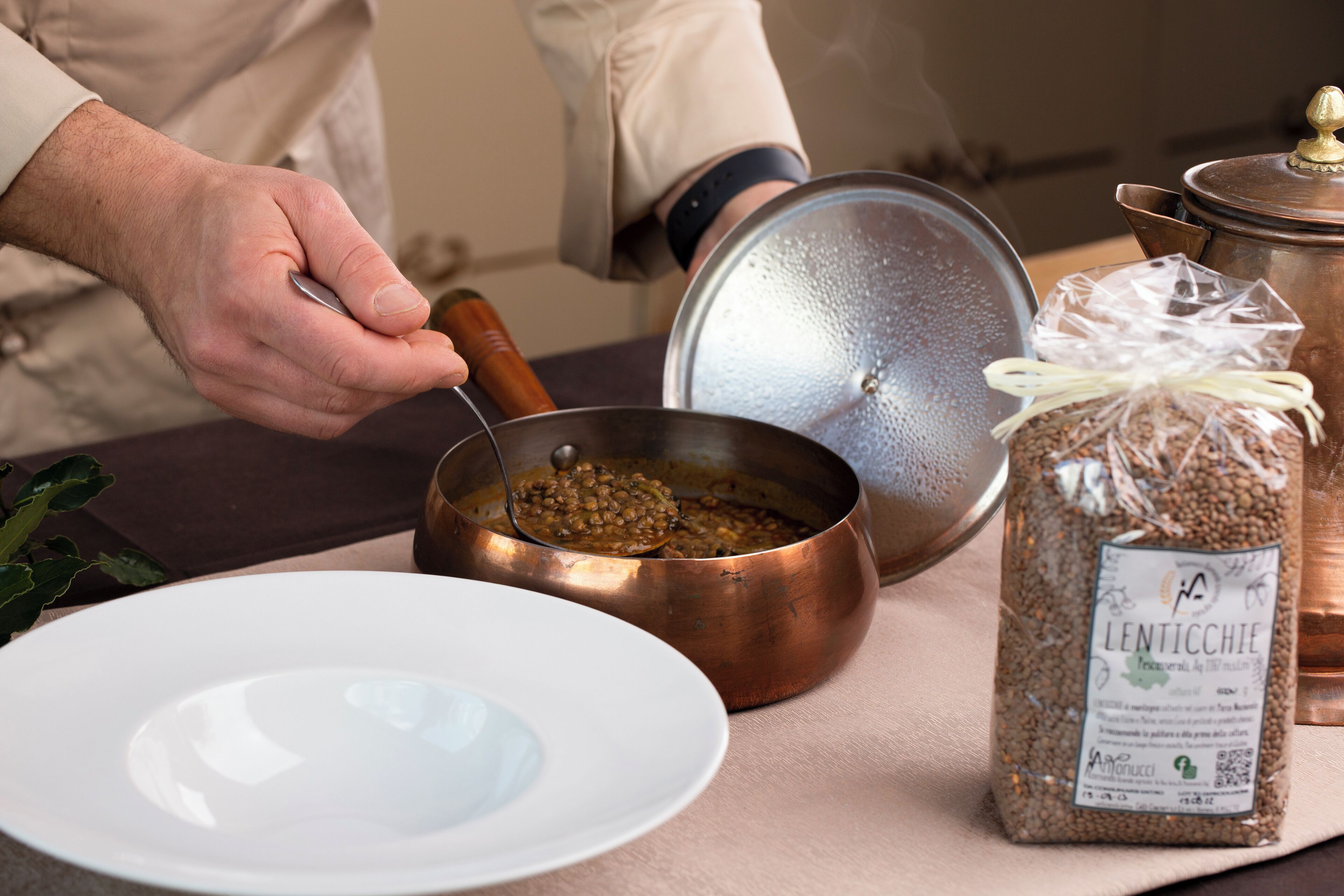 Uma pessoa cozinhando lentilhas em uma panela de cobre, com um prato branco e um saco de lentilhas na mesa.