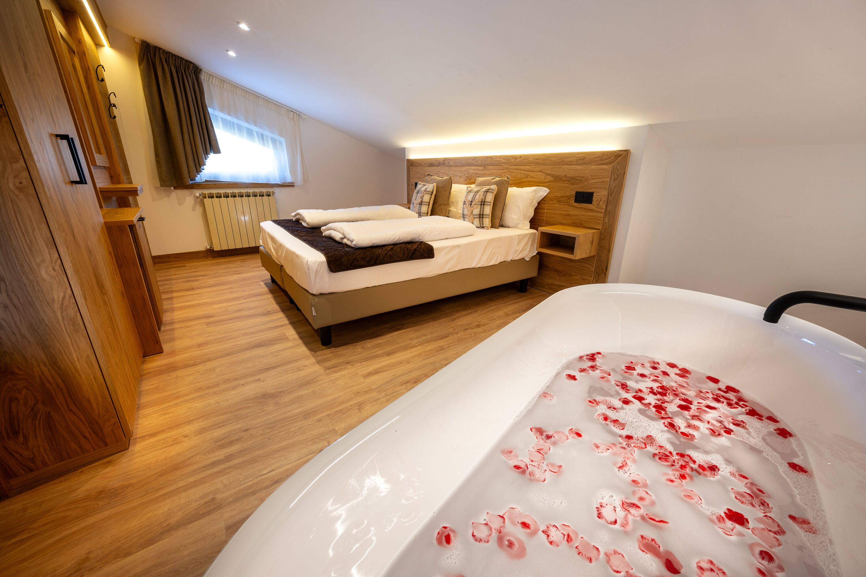 Quarto moderno com cama grande, piso de madeira, armário embutido e banheira com pétalas de rosa.