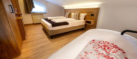 Quarto moderno com cama grande, piso de madeira, armário embutido e banheira com pétalas de rosa.