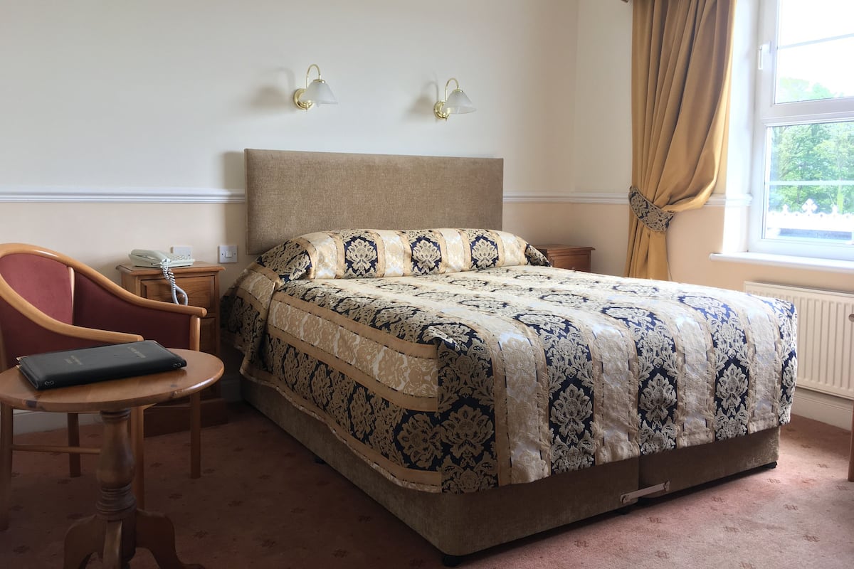 Quarto de hotel com cama, poltrona, mesinha, criado-mudo, abajur e janela com cortinas.