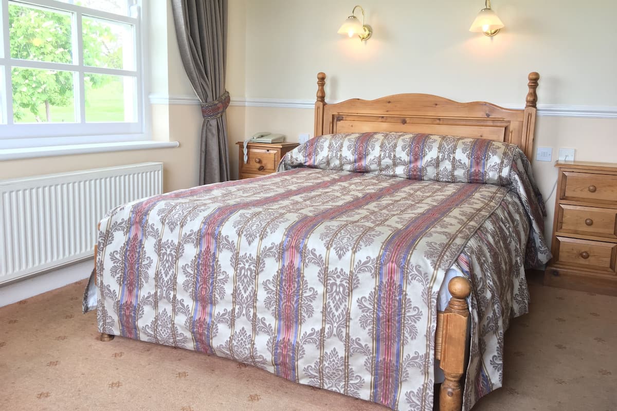 Quarto com cama de madeira, duas mesas de cabeceira, um criado-mudo, uma janela com cortinas e uma luminária na parede.