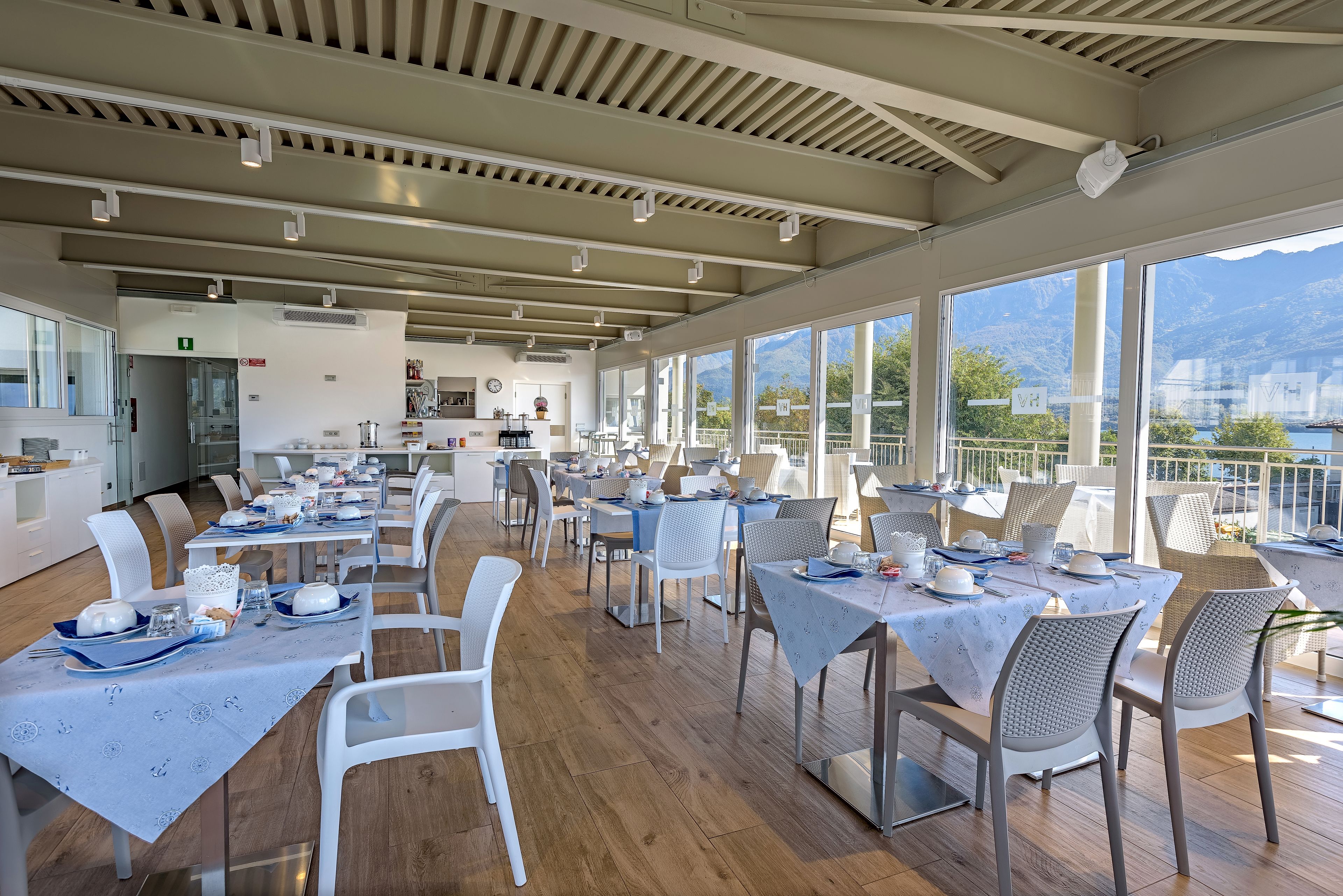 Uma sala de jantar espaçosa com mesas preparadas para vários convidados, amplas janelas com vista para as montanhas, e uma área de cozinha ao fundo.