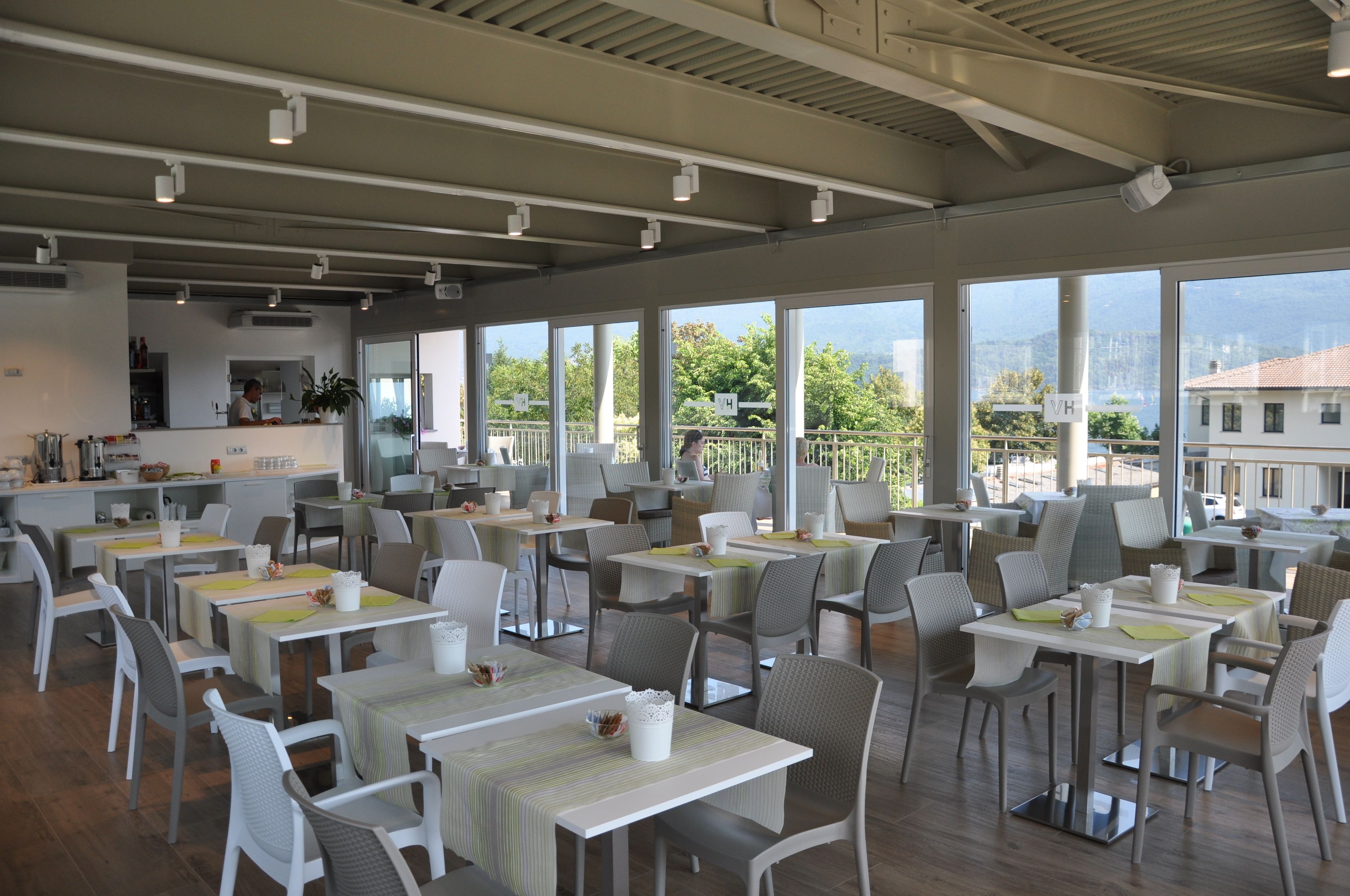 Um restaurante espaçoso com várias mesas e cadeiras, grandes janelas com vista para a cidade e uma área de cozinha visível ao fundo.