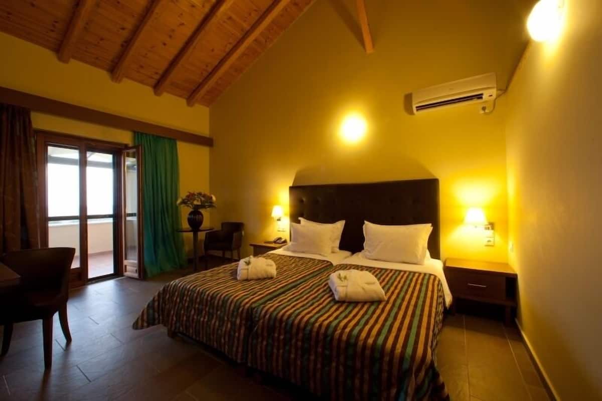 Quarto de hotel com uma cama grande, duas luminárias de cabeceira, uma mesa pequena, uma cadeira e uma porta de vidro deslizante para uma área externa.