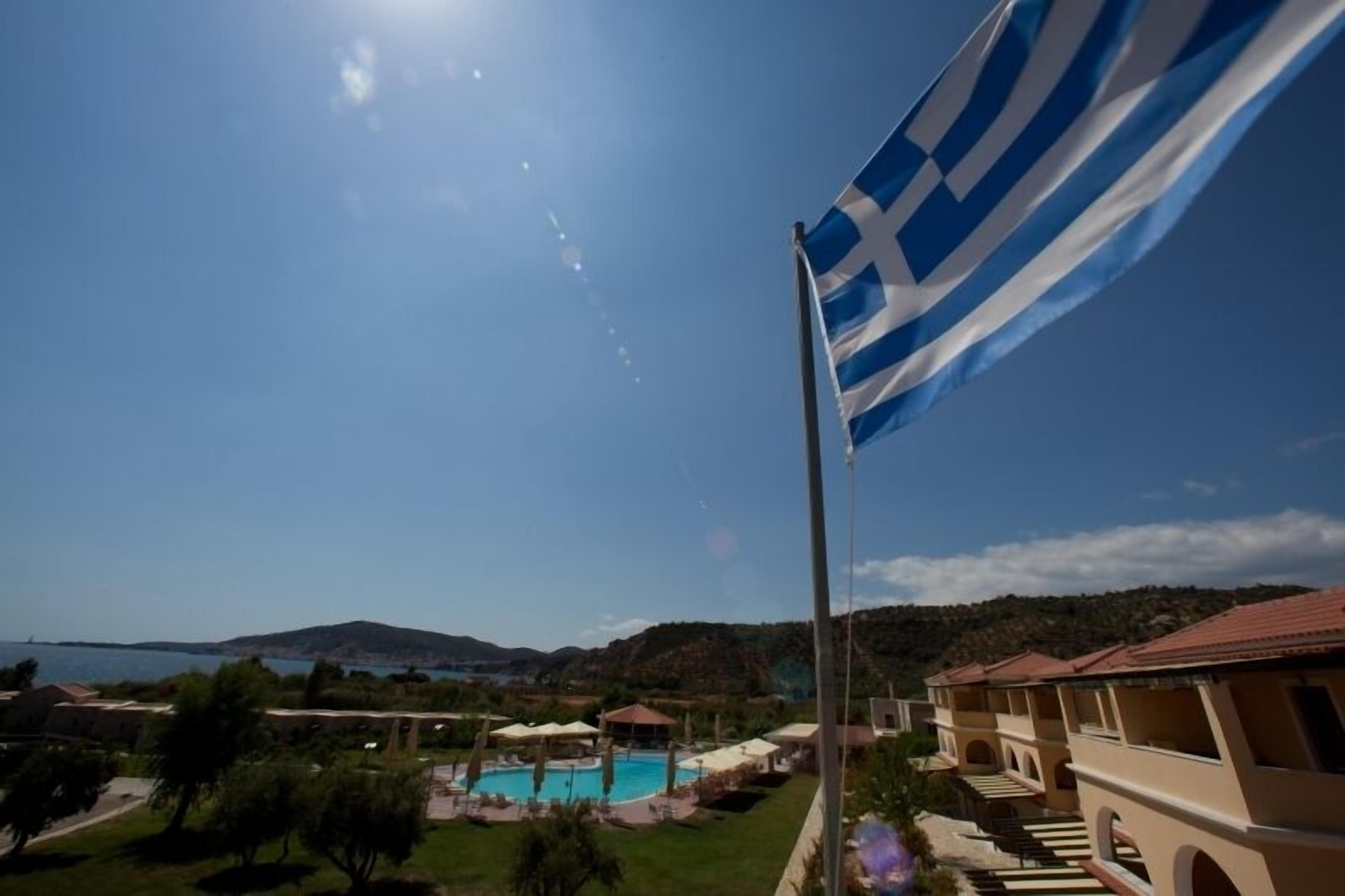 Um mastro com uma bandeira, piscina e edifícios ao fundo.