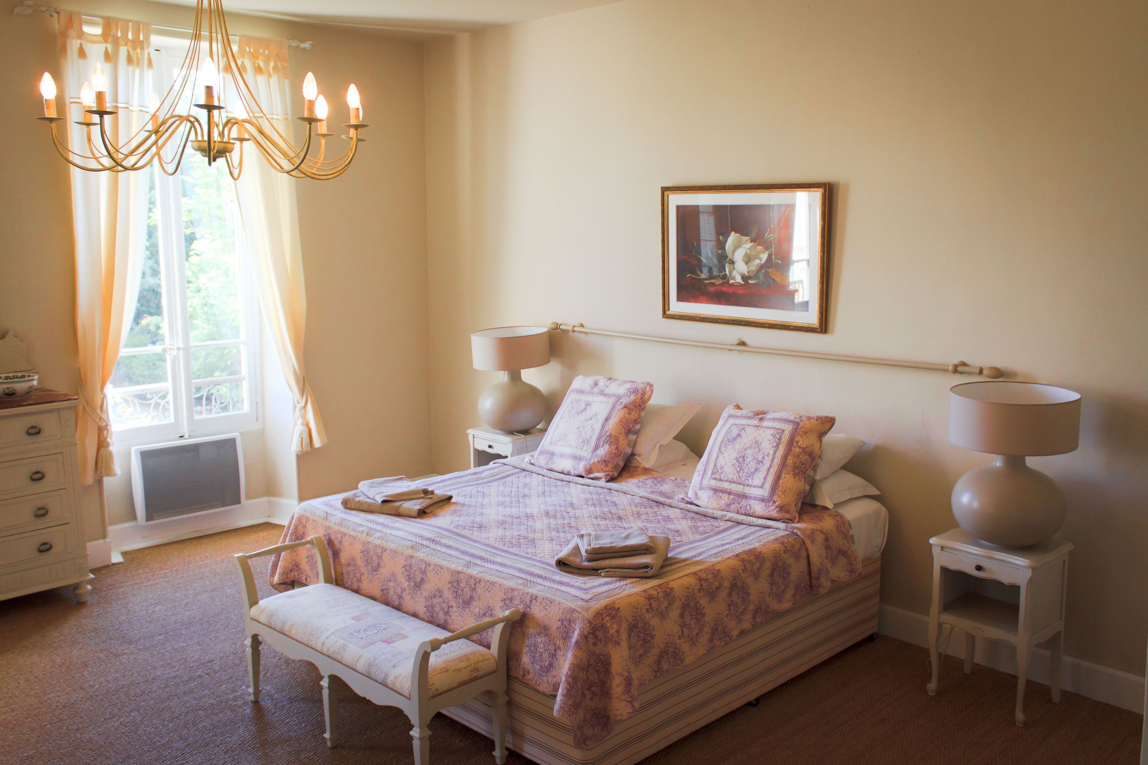 Quarto com cama, mesas de cabeceira, uma cadeira, um candelabro, janela com cortinas e um quadro na parede.