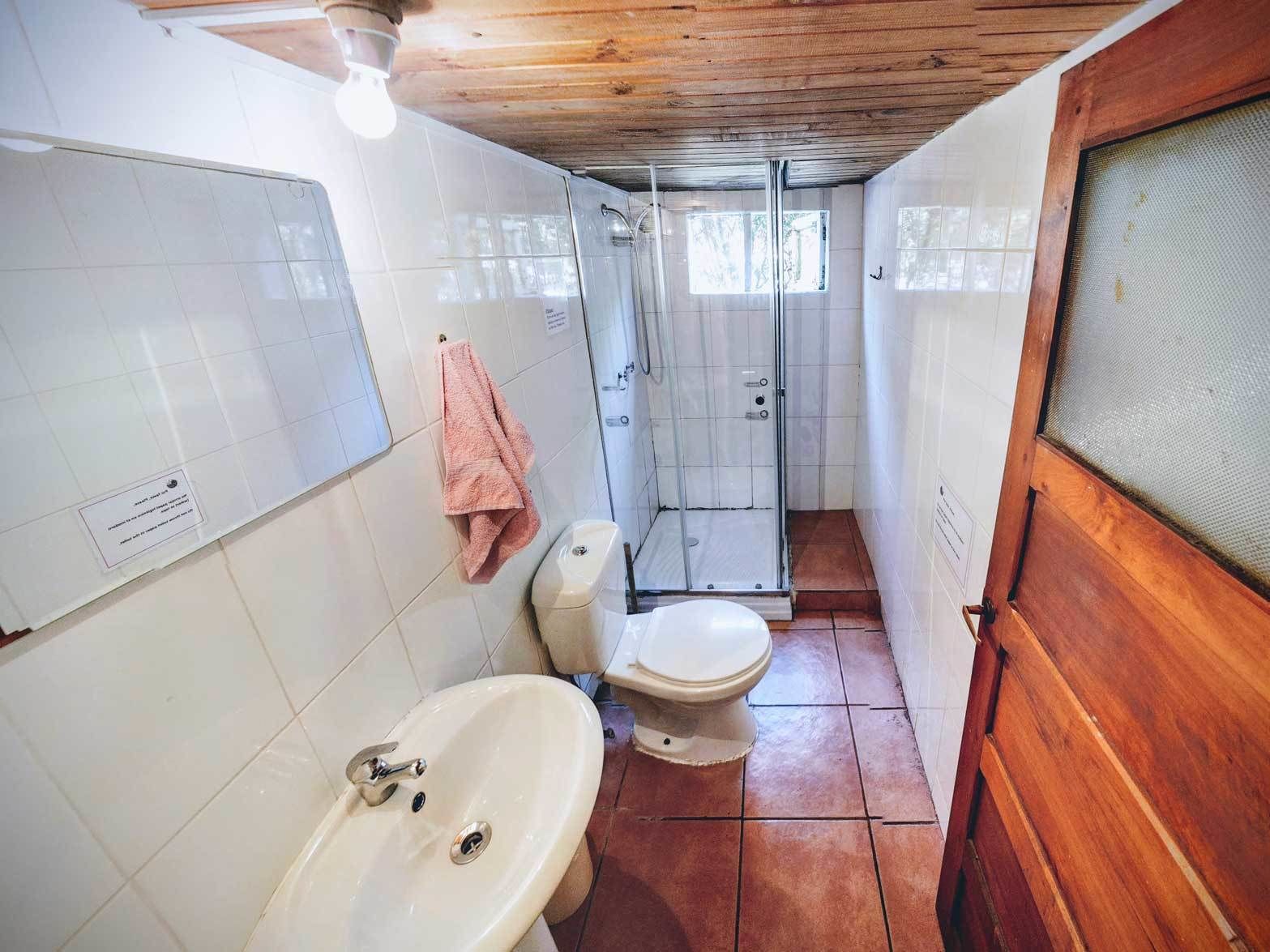 Casa de banho com teto de madeira, paredes em azulejo branco, toalha rosa, lavatório, sanita e cabine de duche em vidro.