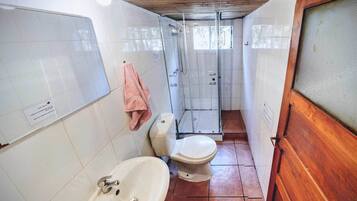 Casa de banho com teto de madeira, paredes em azulejo branco, toalha rosa, lavatório, sanita e cabine de duche em vidro.