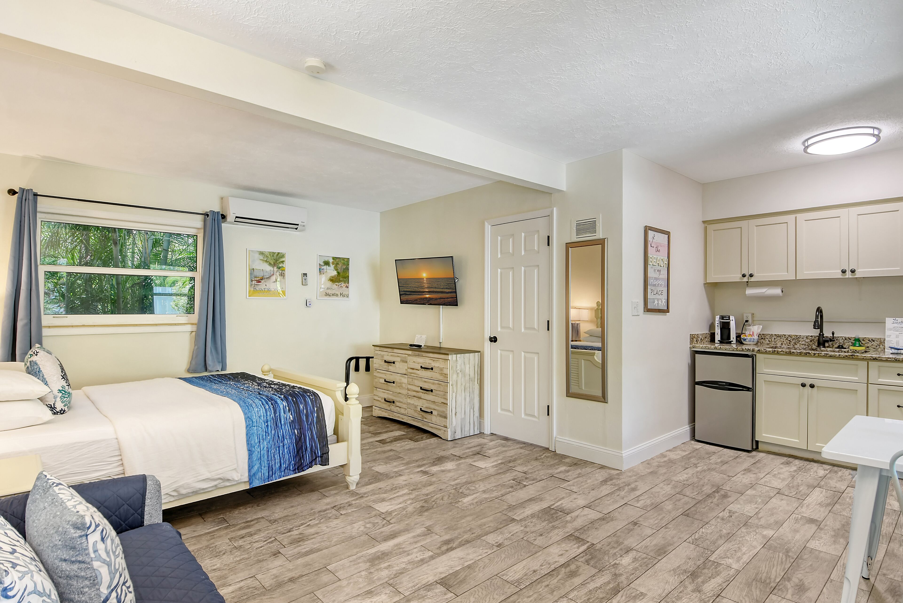 Quarto compacto com cama, escrivaninha, cozinha compacta e porta que dá para outro espaço.