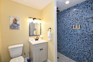 Casa de banho com sanita, lavatório e espelho. Inclui chuveiro com azulejos de mosaico azul.