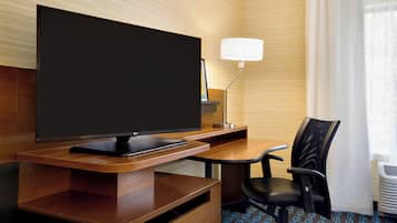 Quarto de hotel com televisão de tela plana sobre uma escrivaninha de madeira, uma cadeira de escritório preta, uma luminária e uma janela com cortinas.