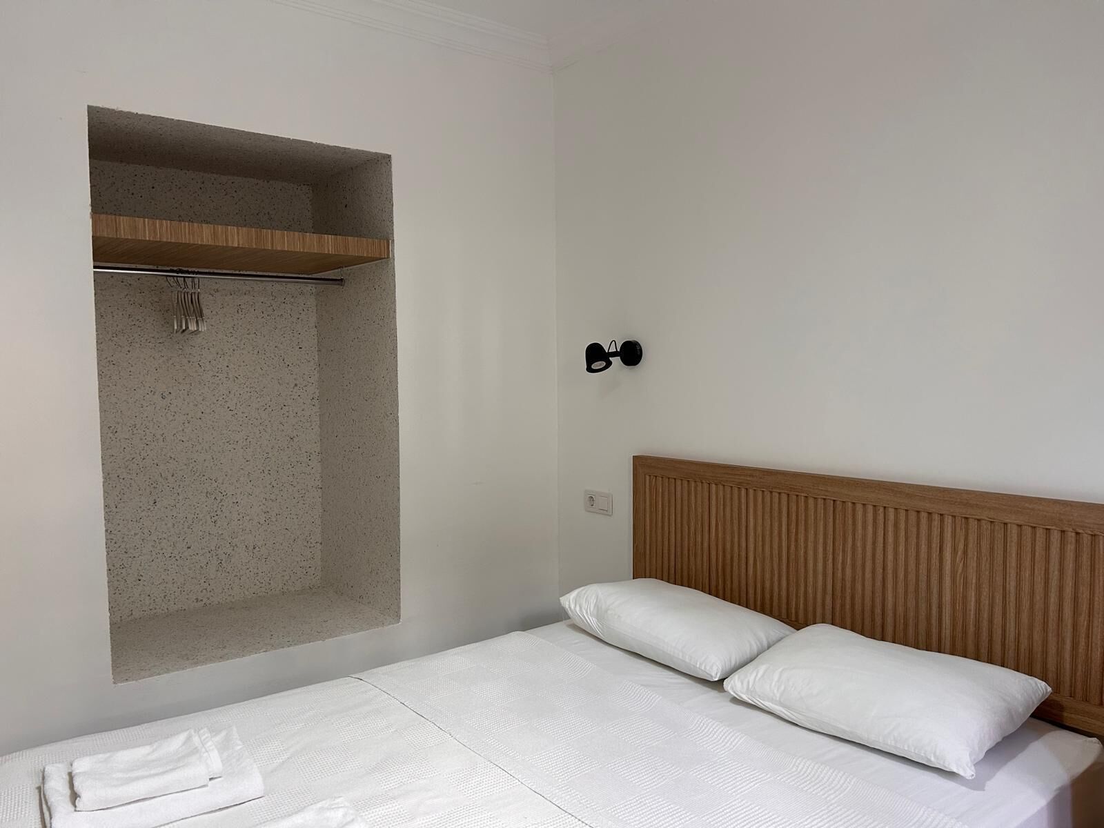 Quarto com cama, cabeceira de madeira, área de chuveiro e luminária fixada na parede.
