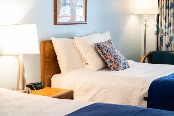 Quarto de hotel com cama, mesa de cabeceira com abajur, relógio e quadro na parede.