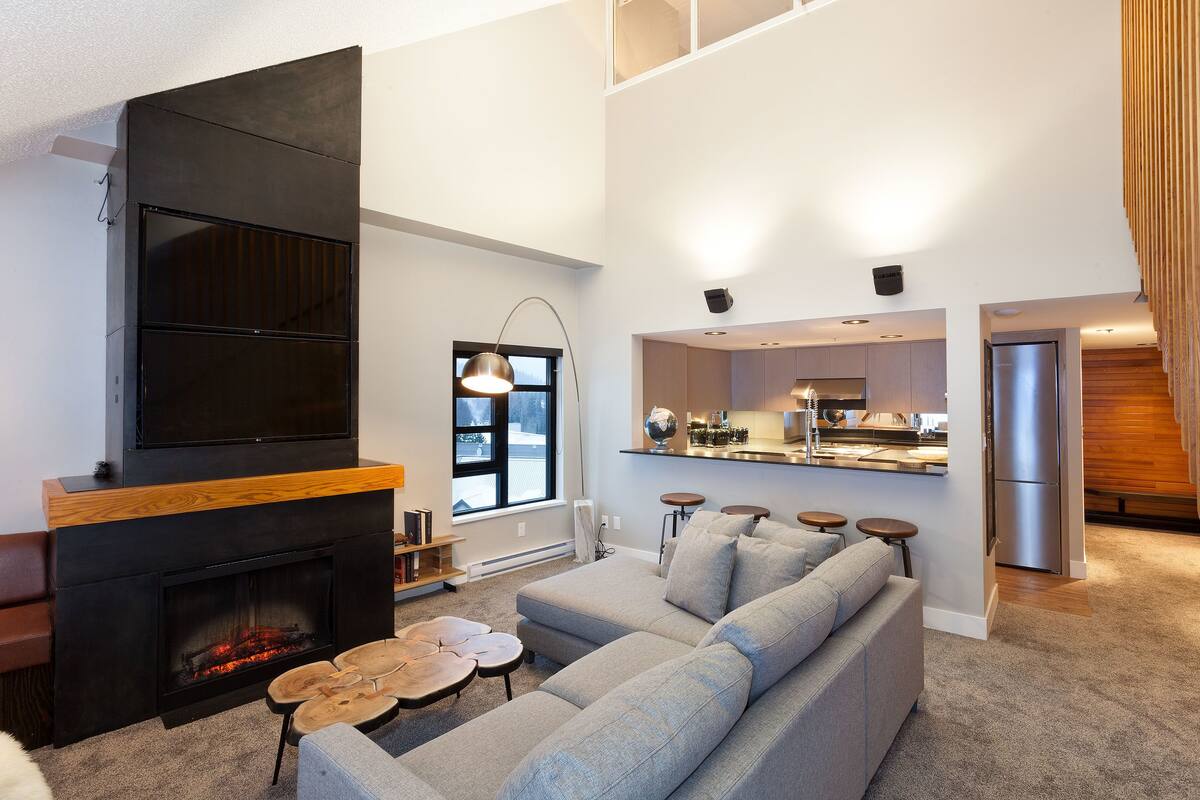 Uma sala de estar moderna com lareira, um amplo sofá modular e uma área de cozinha ao fundo.