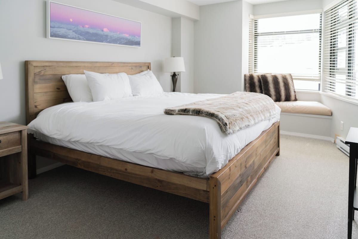Quarto com cama de madeira, roupa de cama branca, mesa de cabeceira, abajur, um banco com manta de pele e janela com persianas.