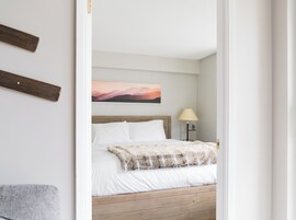 Quarto com cabeceira de madeira, roupa de cama branca, manta de pele, luminária e pintura de montanhas.