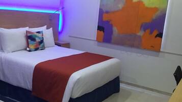 Quarto de hotel com cama, mesa de cabeceira, luminária, quadro e escrivaninha.