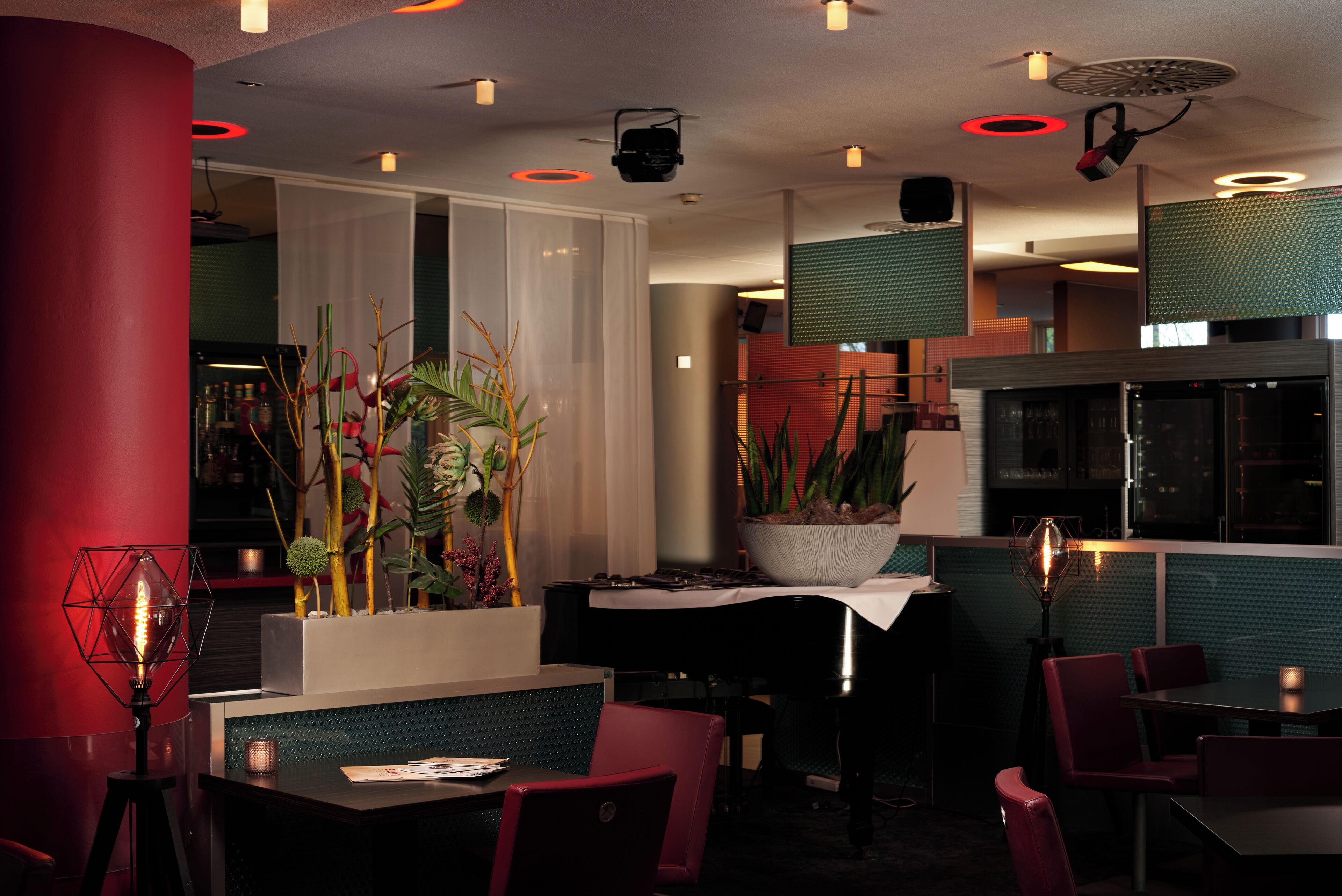 Interior moderno de restaurante com piano, cadeiras vermelhas e diversas plantas.