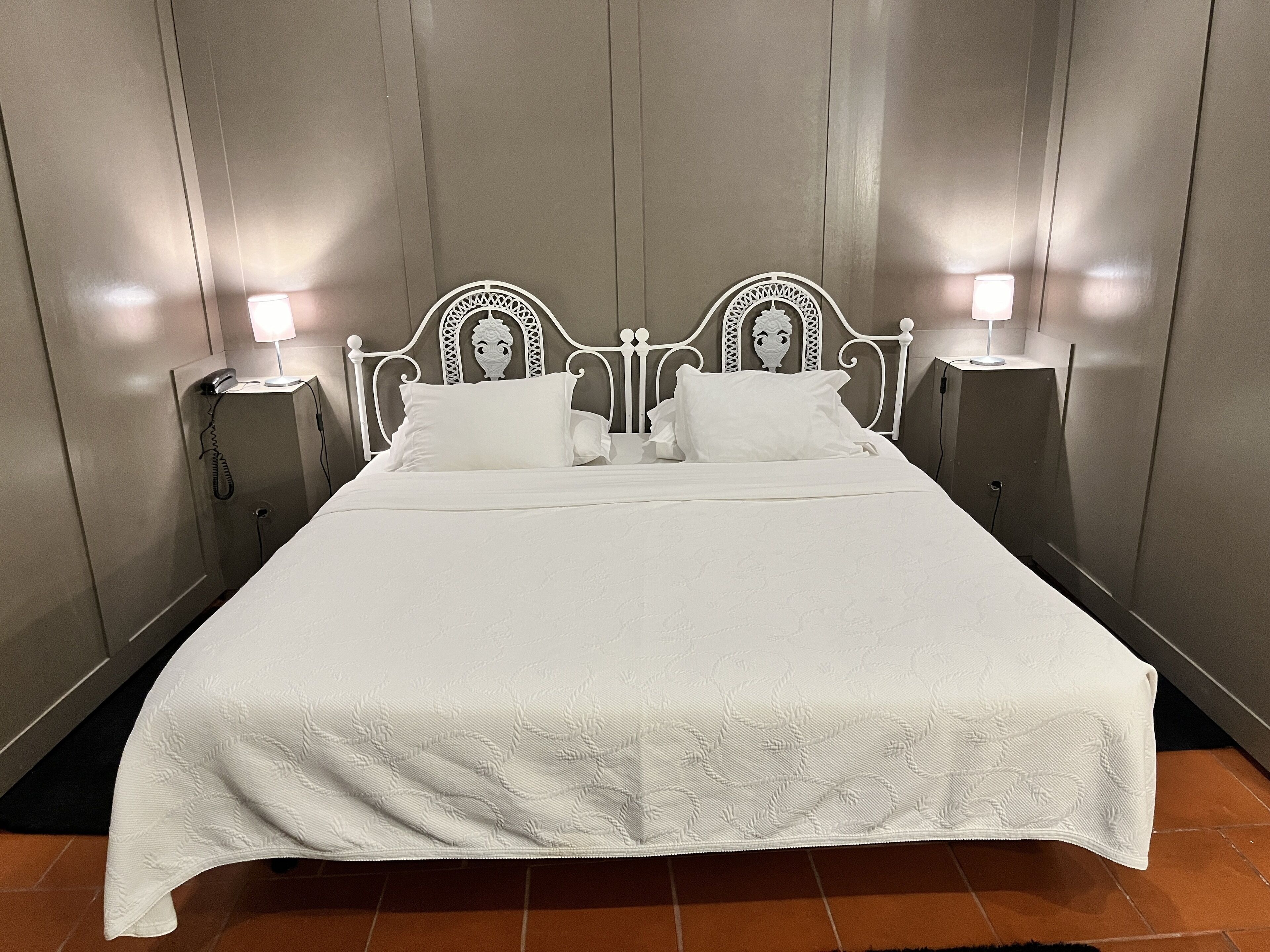 Cama bem arrumada com colcha e travesseiros brancos, acompanhada por duas mesas de cabeceira com abajures, em um quarto com painéis de madeira.