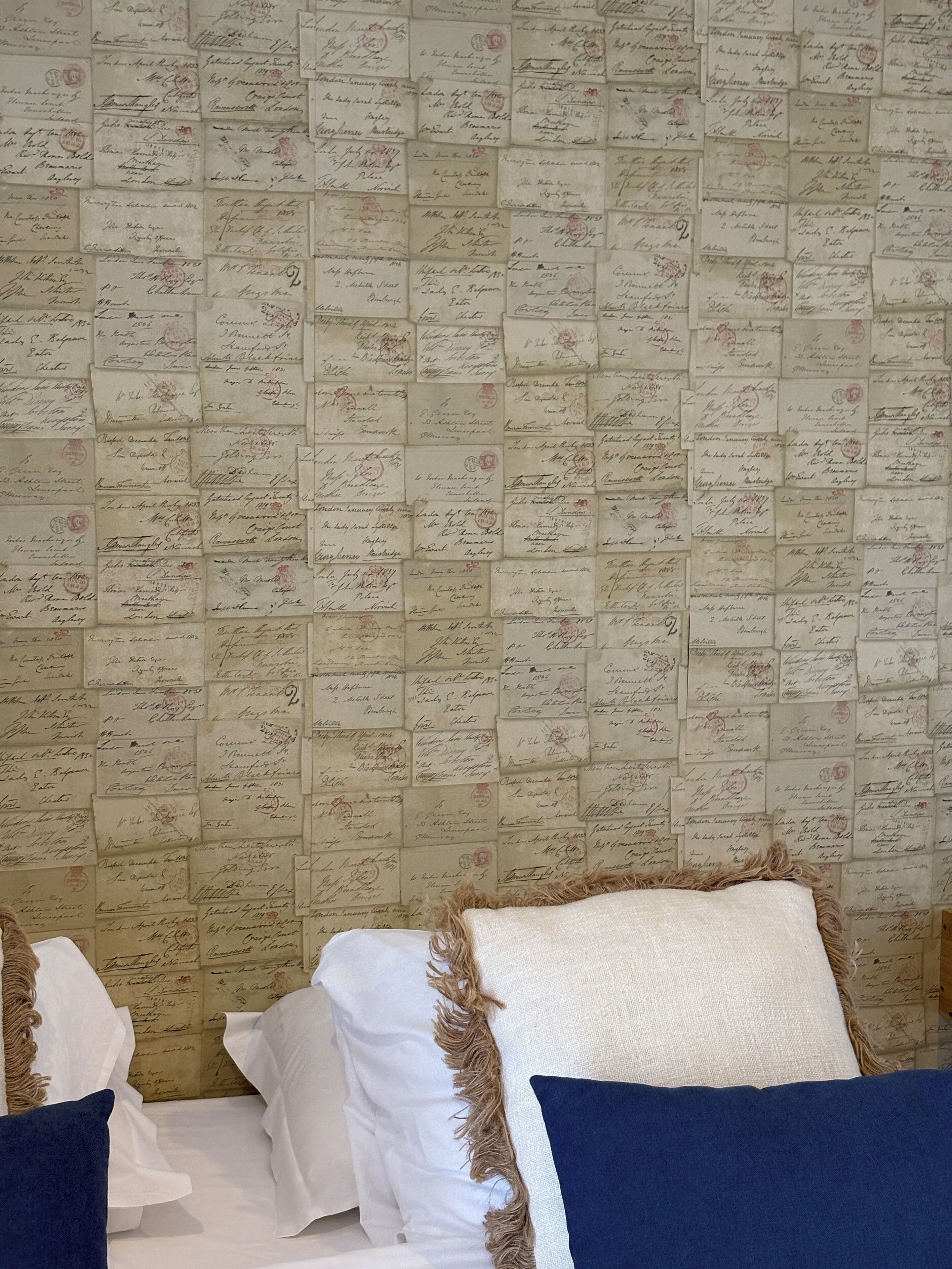 Quarto de hotel com parede coberta por cartões postais antigos, cama com roupa de cama azul e branca, e manta branca.