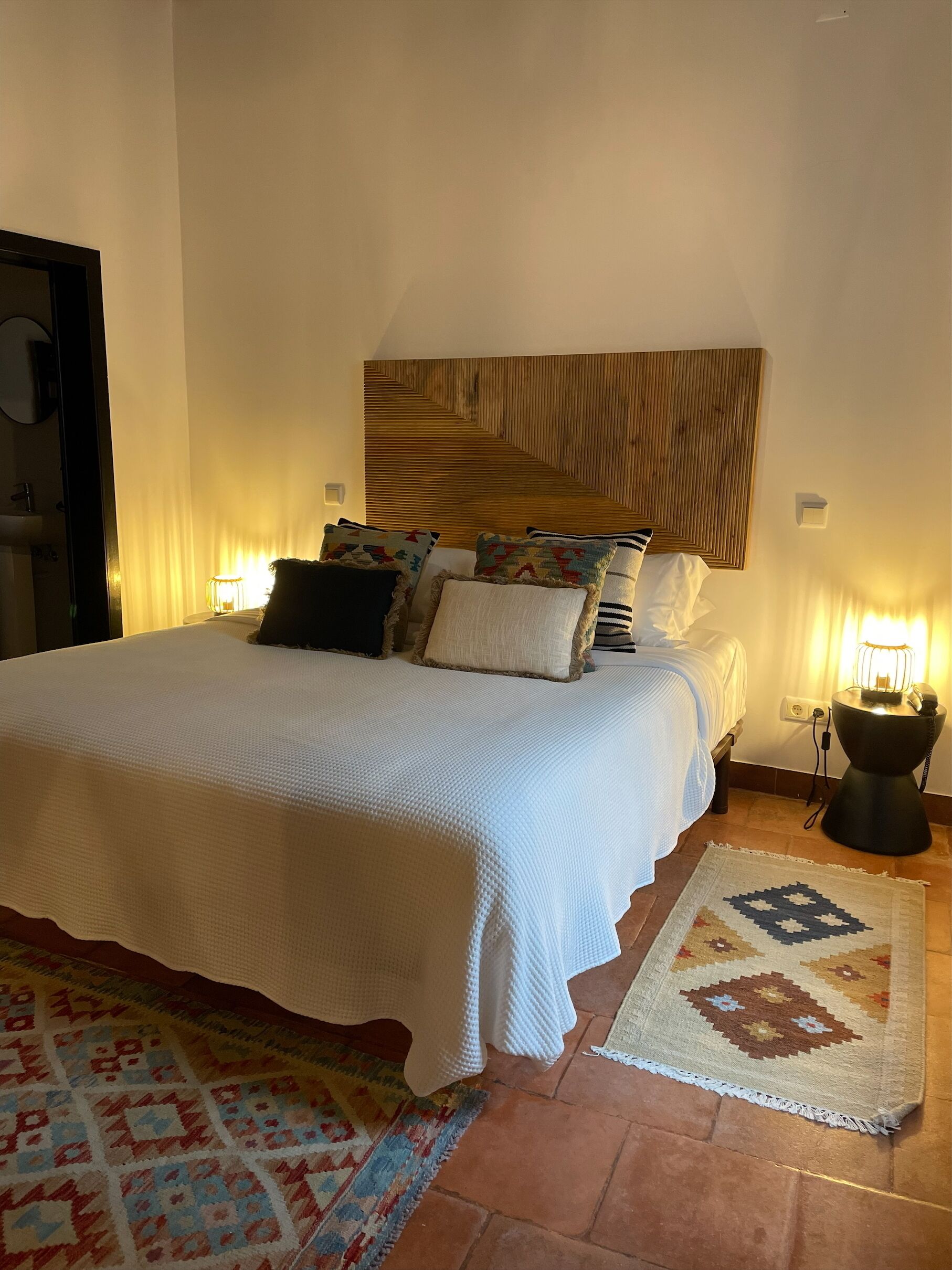 Cama bem arrumada com colcha branca e várias almofadas, cabeceira de madeira, tapete estampado e mesa de cabeceira com abajur.