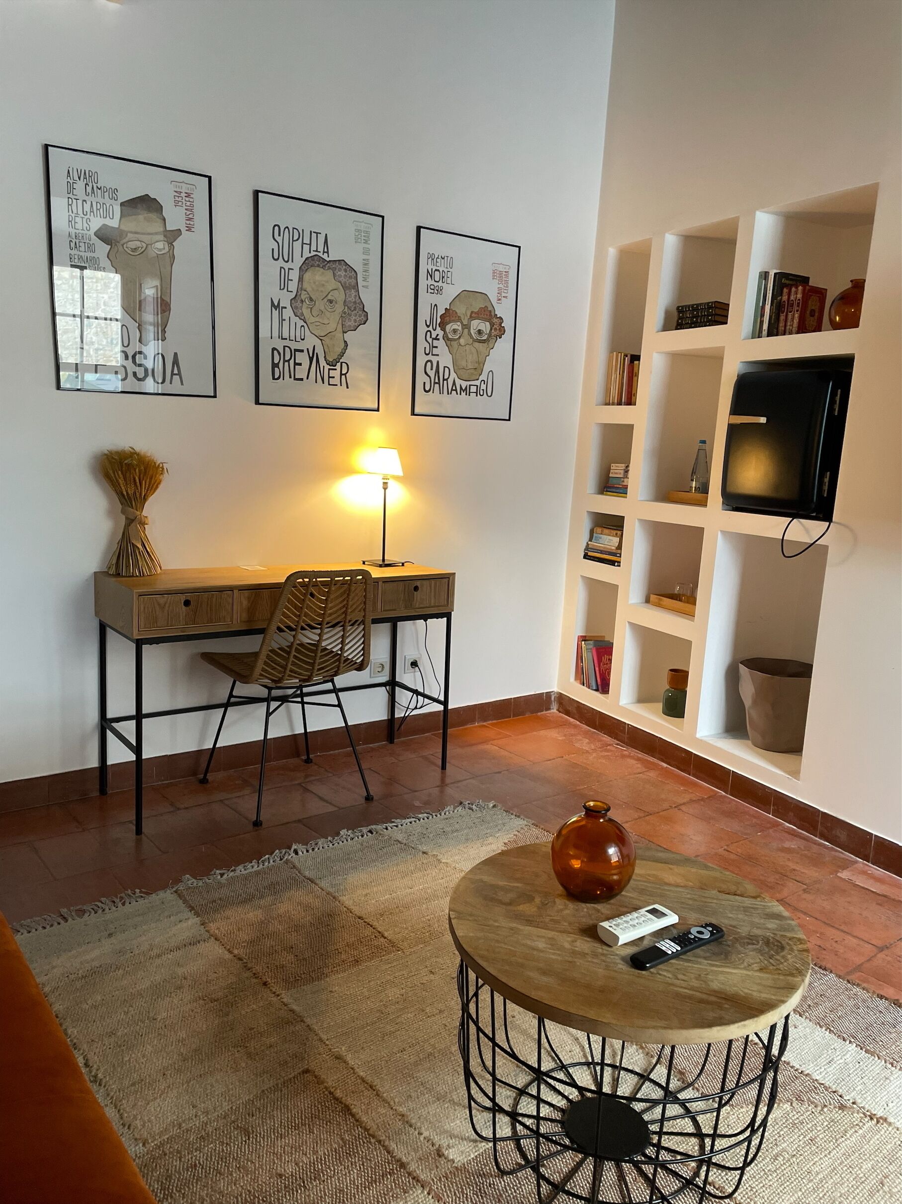 Uma sala de estar acolhedora, com uma escrivaninha de madeira, uma cadeira, uma mesa de centro redonda com um vaso e controles remotos, e uma estante com livros e objetos decorativos.