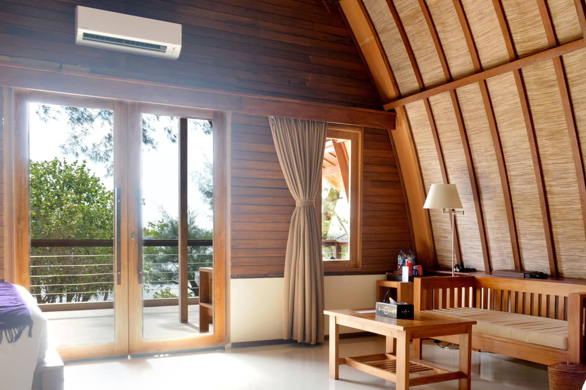 Quarto com teto de madeira, uma cama, uma mesa e um banco. Há uma janela com cortinas e vista para o verde lá fora.
