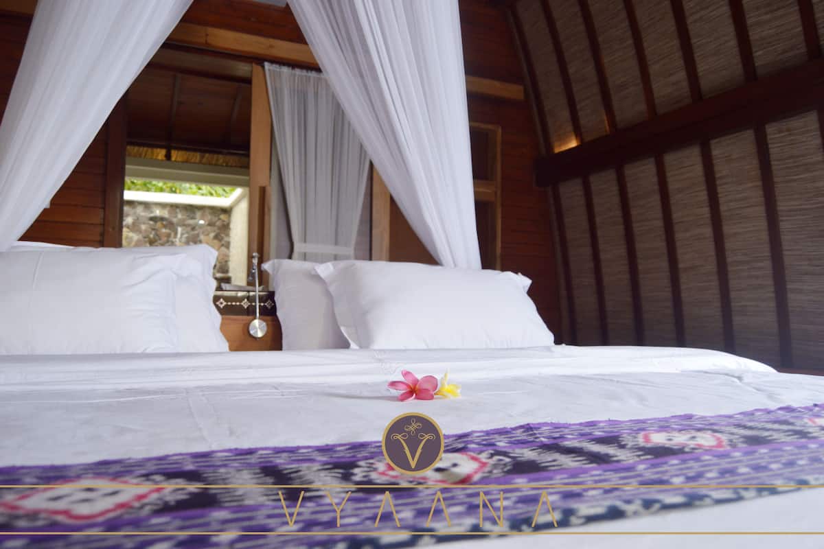 Uma cama com lençóis brancos, um edredom com padrão roxo e branco, e uma flor rosa sobre a cama.