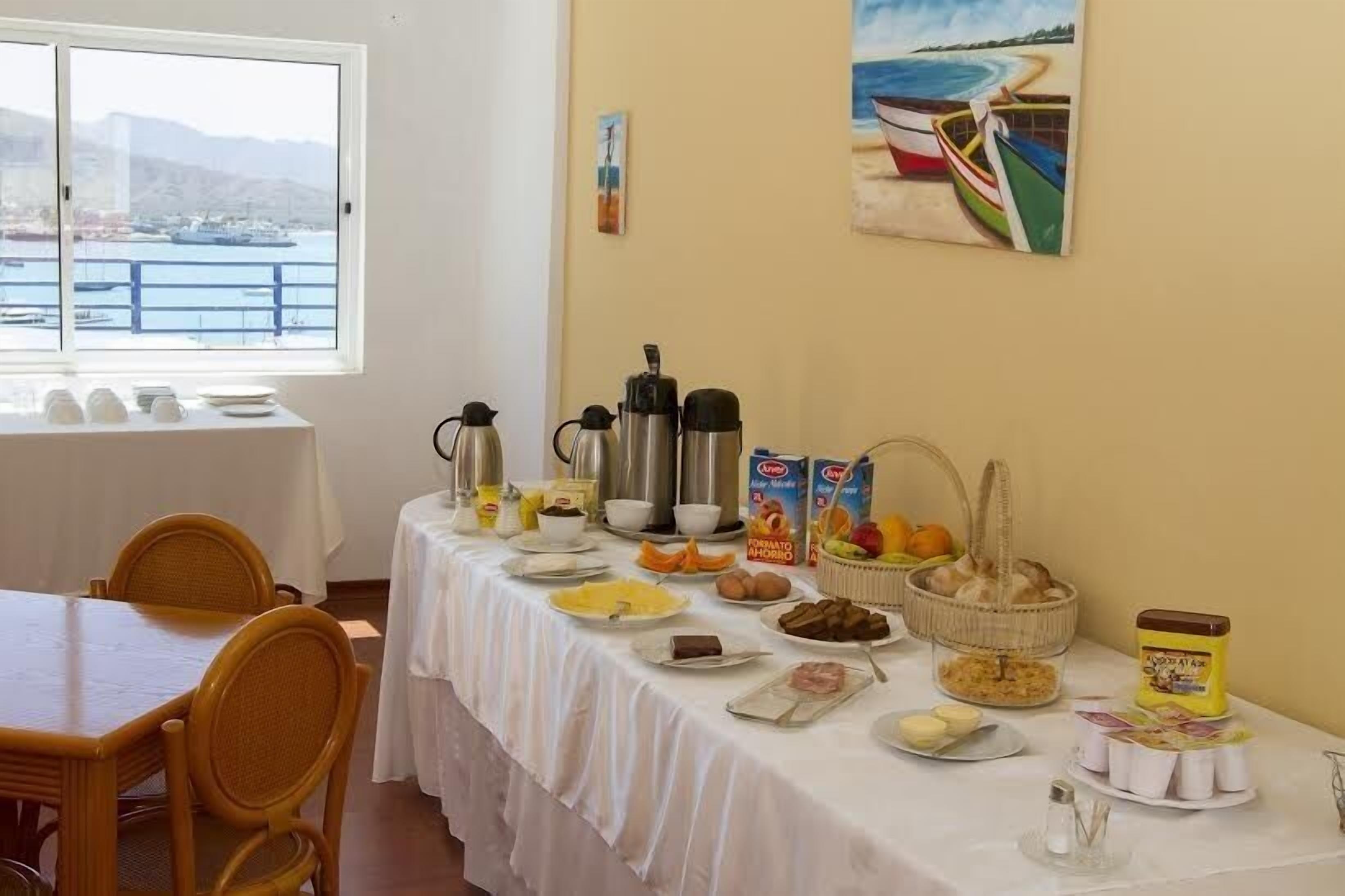 Uma mesa de café da manhã com diversos alimentos, cafeteiras e um quadro de barcos na parede.