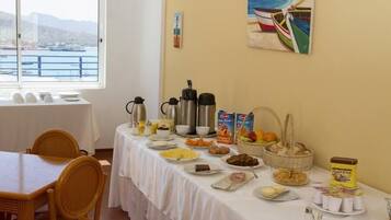 Uma mesa de café da manhã com diversos alimentos, cafeteiras e um quadro de barcos na parede.