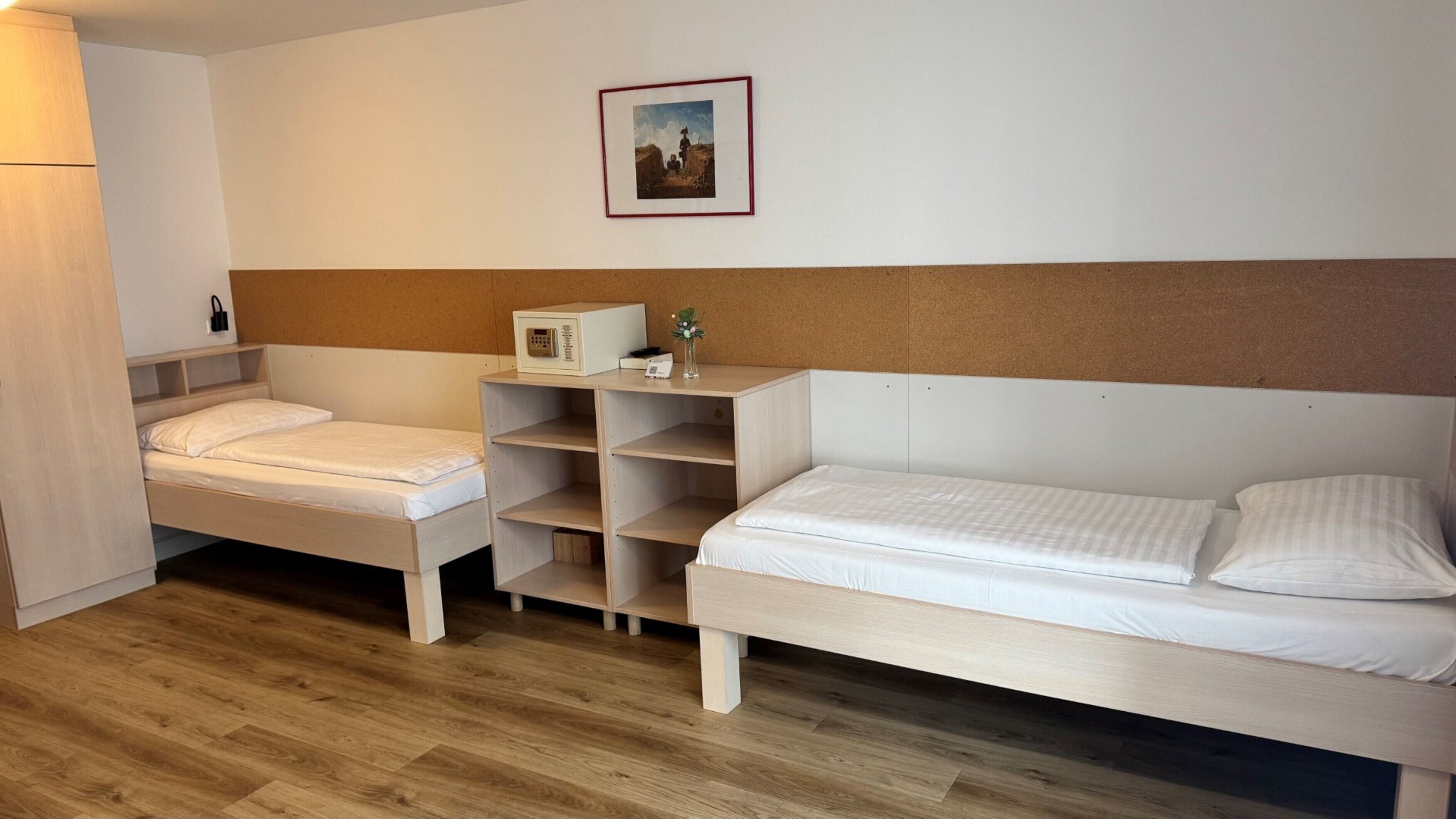 Quarto com duas camas, uma mesa com computador, uma mesinha com um vaso e um quadro na parede.