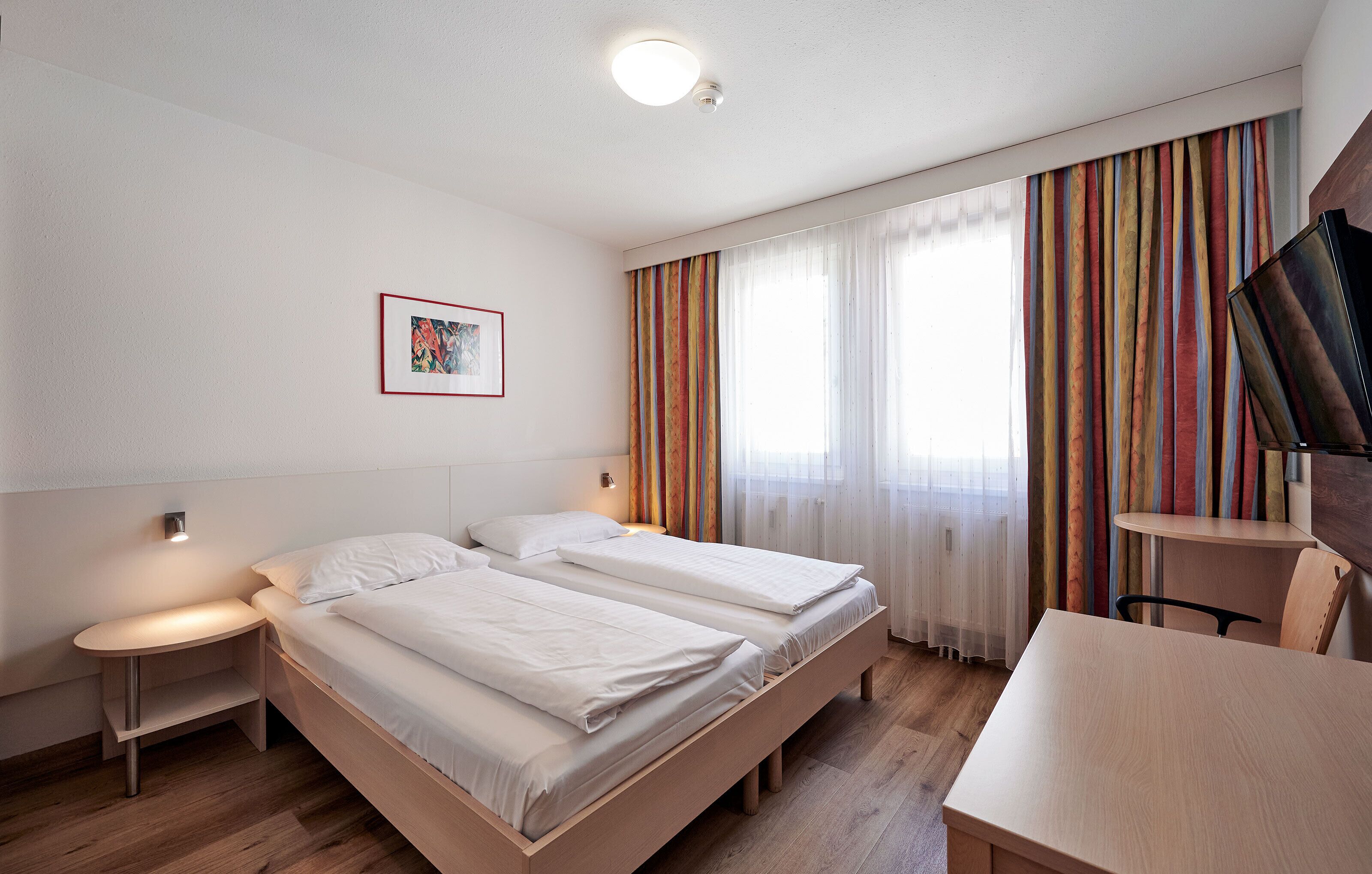 Quarto de hotel com duas camas, uma escrivaninha, televisão e um quadro na parede.