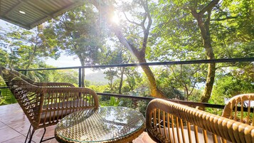 Uma mesa com tampo de vidro e cadeiras de vime numa varanda com vista para uma paisagem verdejante.