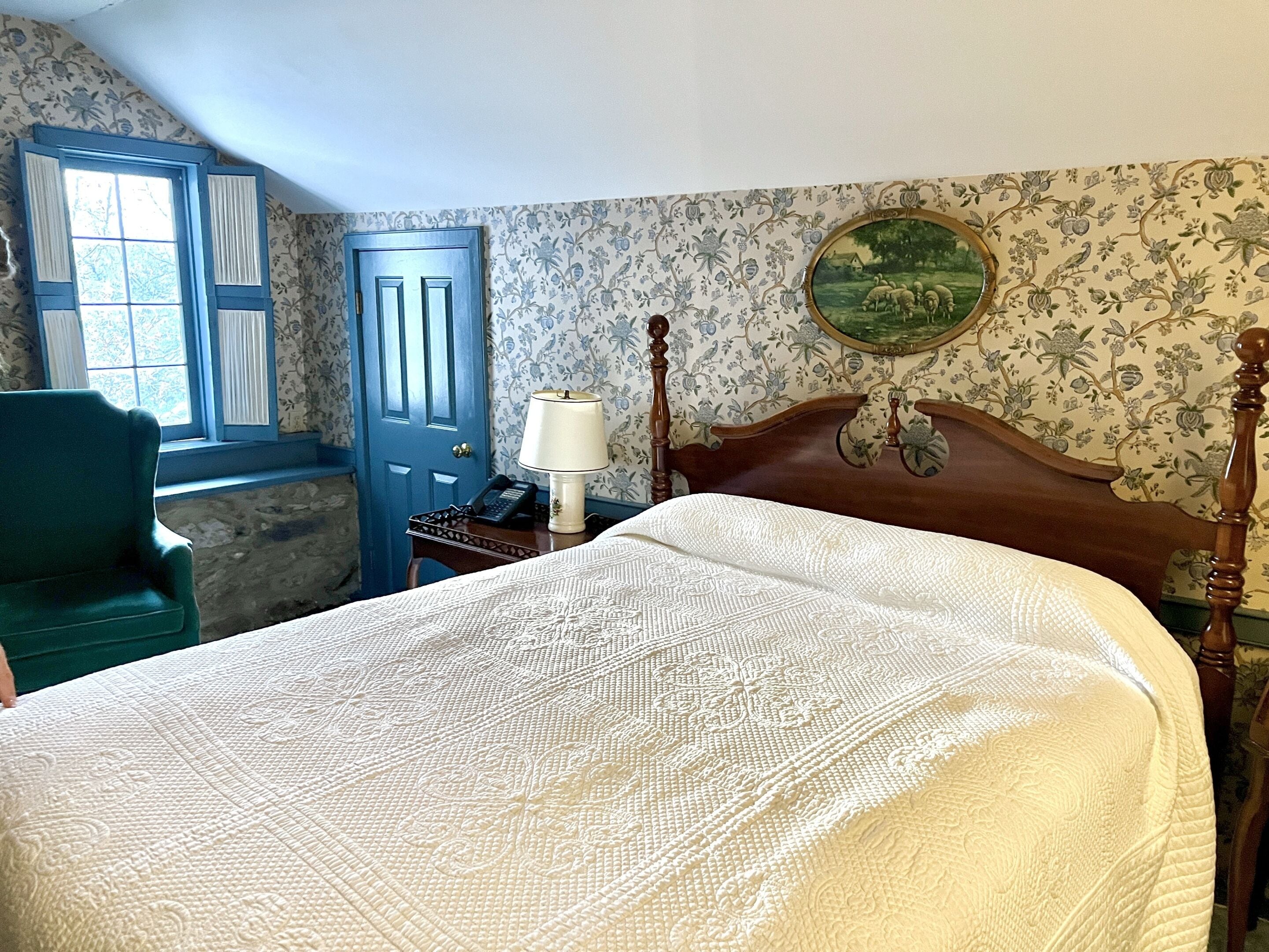 Quarto com cama grande, cabeceira de madeira, mesa de cabeceira com abajur, telefone e uma cadeira.