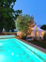Piscina com deck ladrilhado, rodeada por árvores e uma área coberta com luzes de corda.